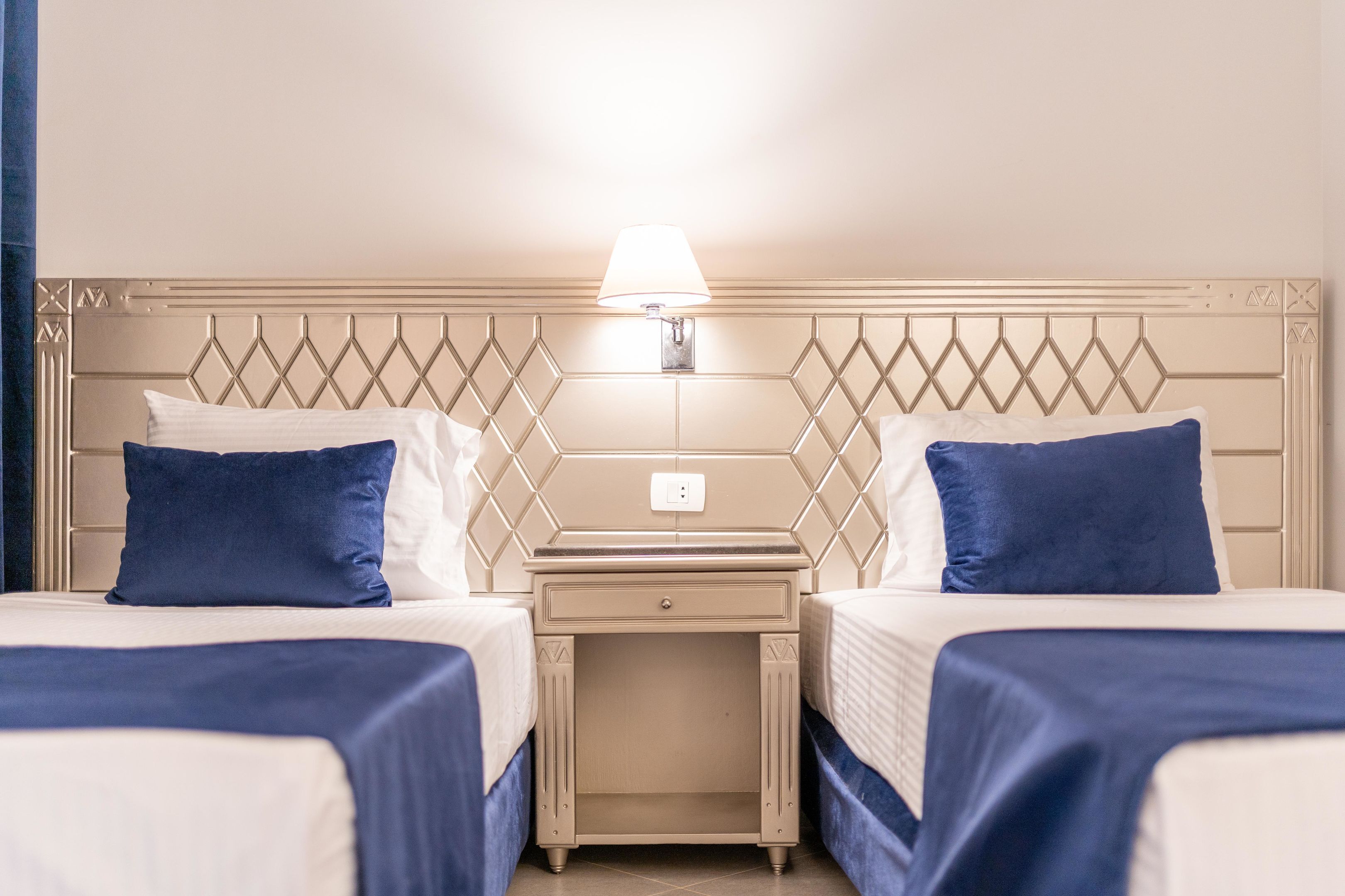 Quarto de hotel com duas camas, cada uma com colcha azul e almofadas brancas. Há uma mesinha de cabeceira entre as camas, com uma luminária e uma gaveta.
