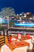Mesa de jantar posta com toalha de padrão, guardanapos vermelhos e copos, com vista para uma piscina e edifícios à noite.