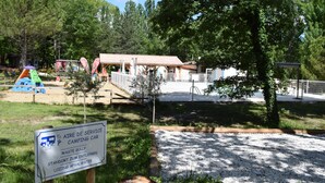 Uma área de camping com um pavilhão coberto, um parquinho e uma placa indicando um ponto de recolha de lixo.