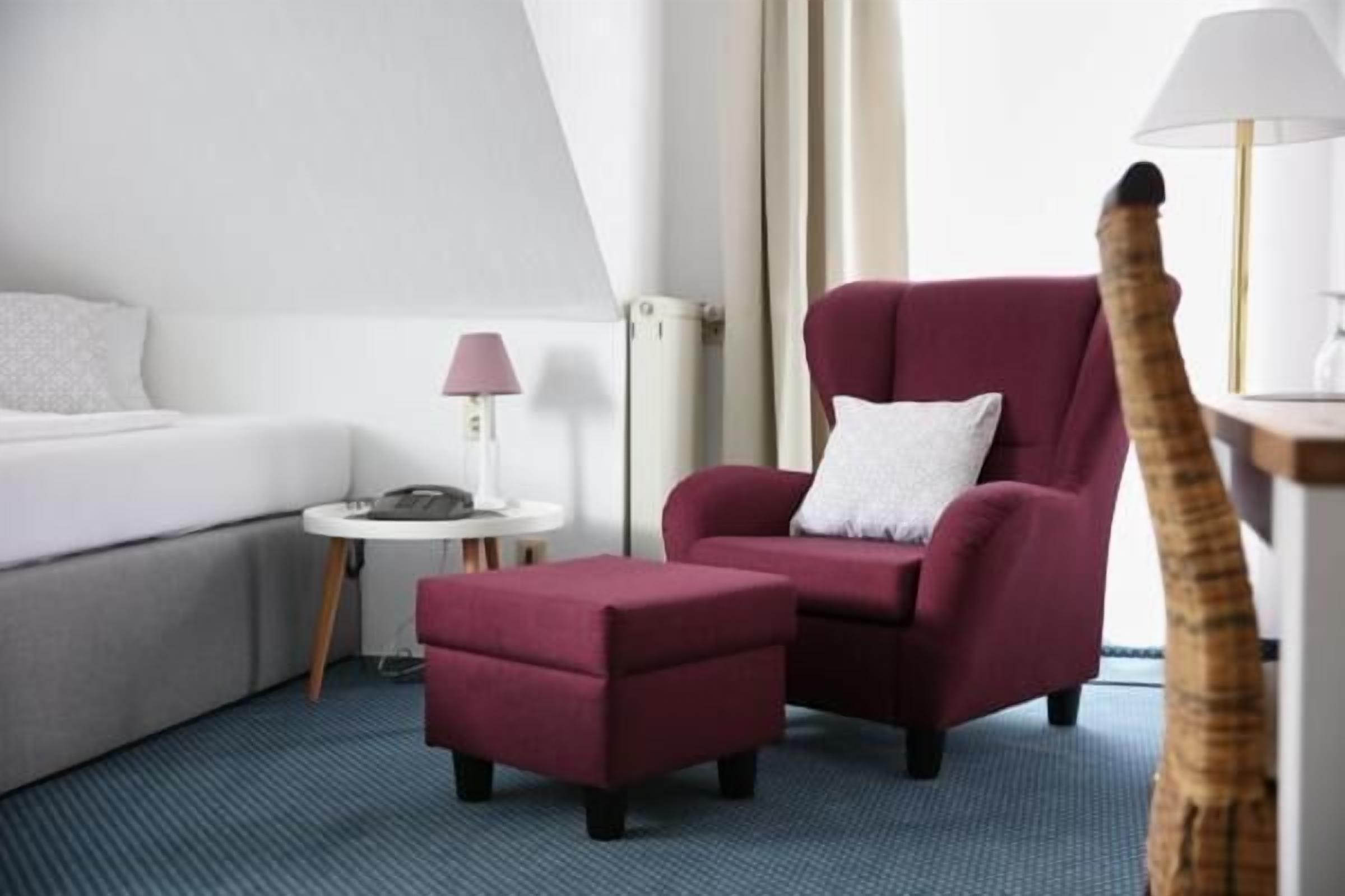 Quarto de hotel com uma cama, uma poltrona vermelha, um banquinho combinado, uma mesinha redonda com abajur e uma janela grande com cortinas.