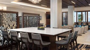 Uma sala de jantar moderna com uma grande mesa retangular rodeada por cadeiras estofadas, um lustre decorativo e vista para o exterior através de grandes janelas.