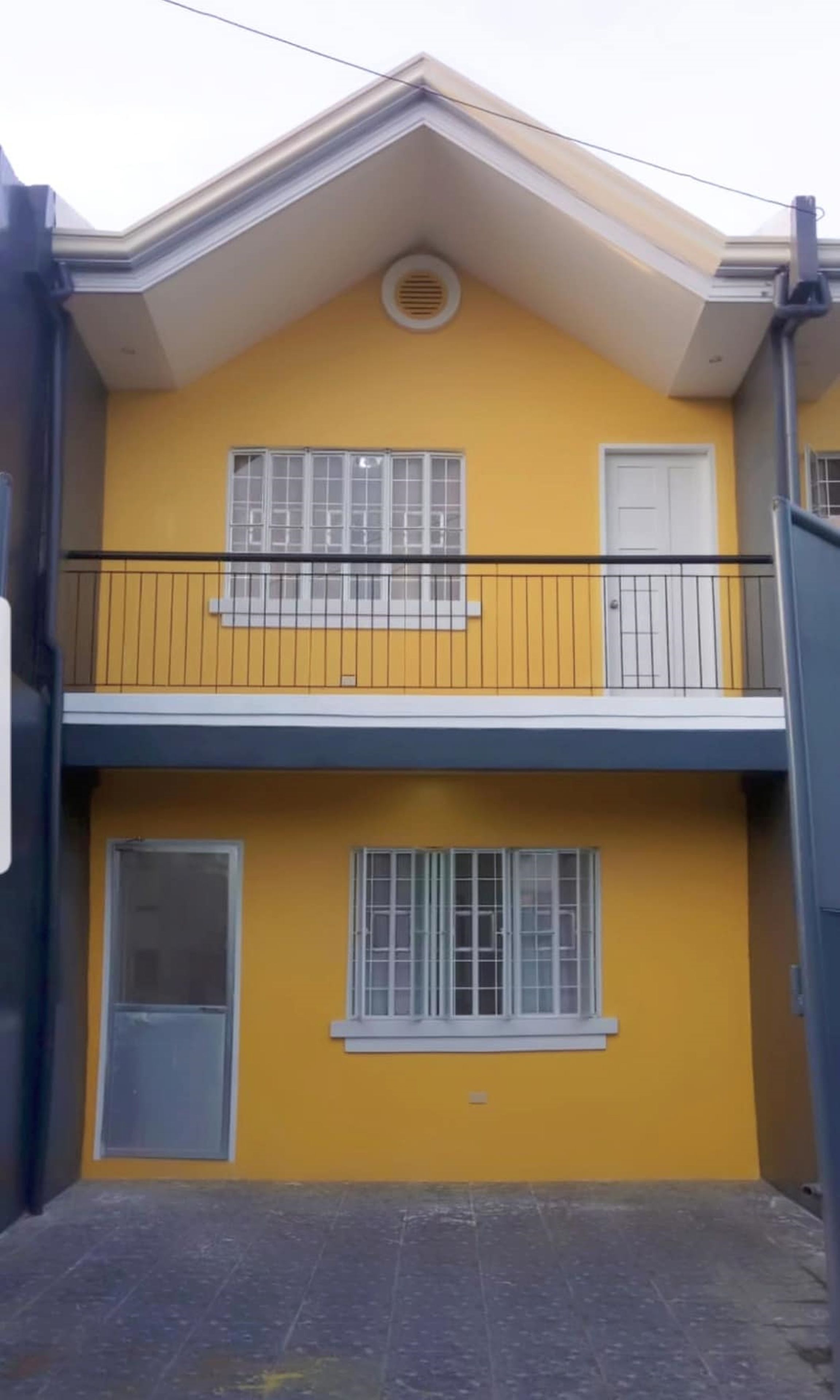 Um edifício de dois andares com fachada amarela, varanda no andar superior e entrada protegida por portão.