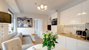 Cozinha moderna com armários brancos, área de refeições com mesa e cadeiras brancas, e sala de estar com sofá e mesa de centro.