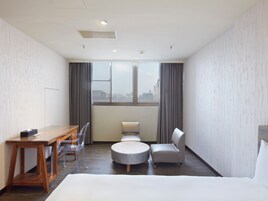 Quarto de hotel com cama, escrivaninha, cadeira, mesa redonda e vista da cidade pela janela.