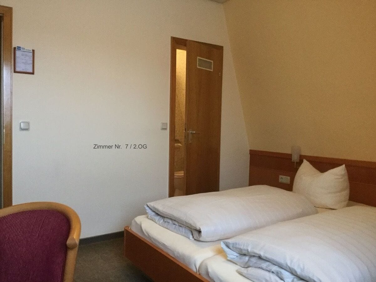 Quarto de hotel com duas camas, uma cabeceira de madeira, uma cadeira vermelha e uma porta com um aviso.