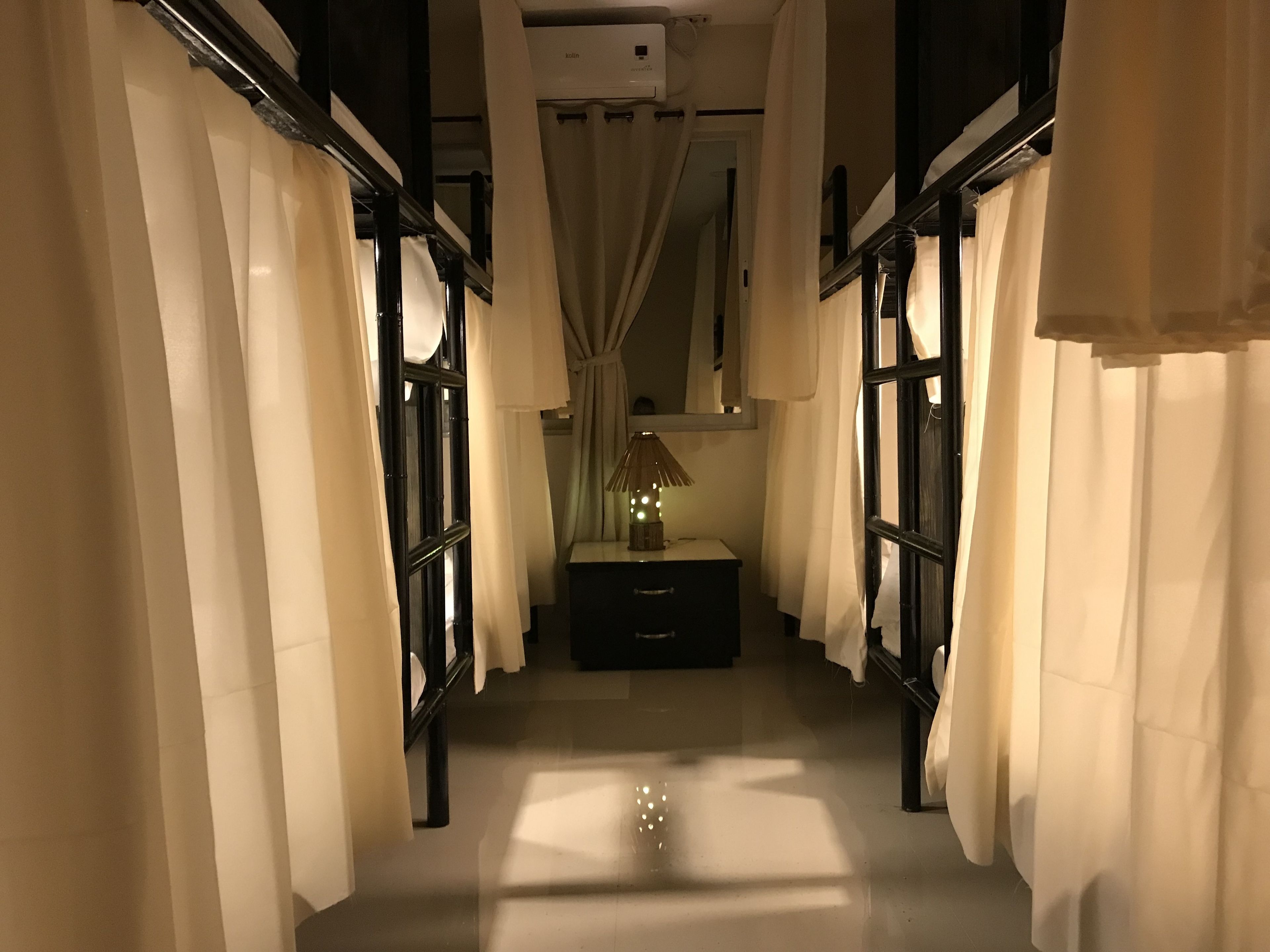 Um corredor estreito com cortinas brancas em ambos os lados, uma pequena mesa com abajur e um espelho na parede.