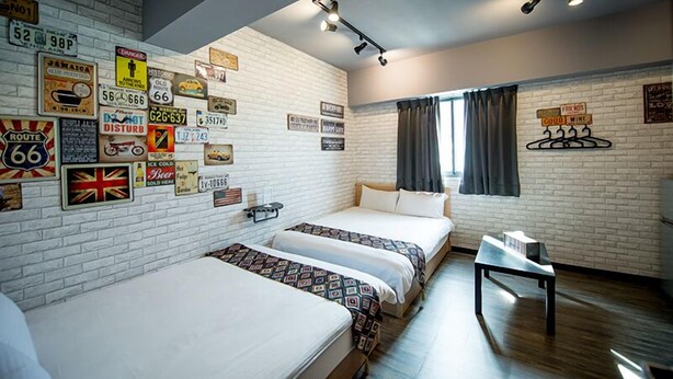 Quarto de hotel com três camas, parede de tijolos com vários avisos, janela com cortinas e uma pequena mesa.