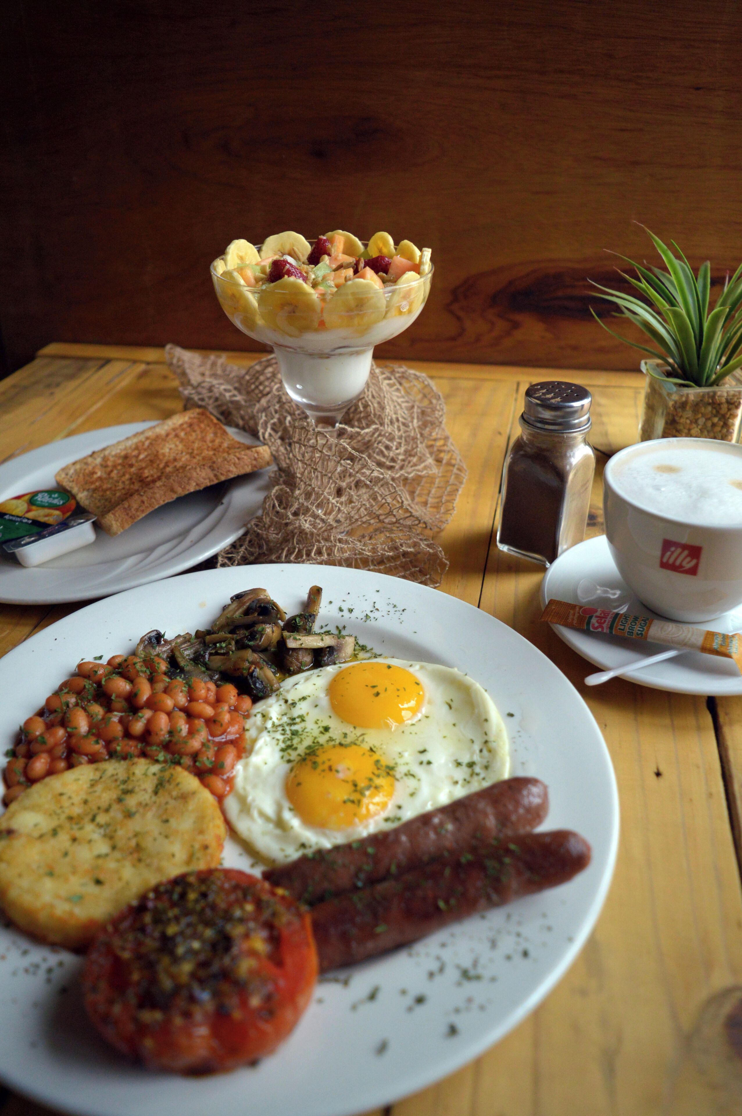 Prato de café da manhã com ovos, linguiça, feijão, cogumelos e uma saladeira de frutas.