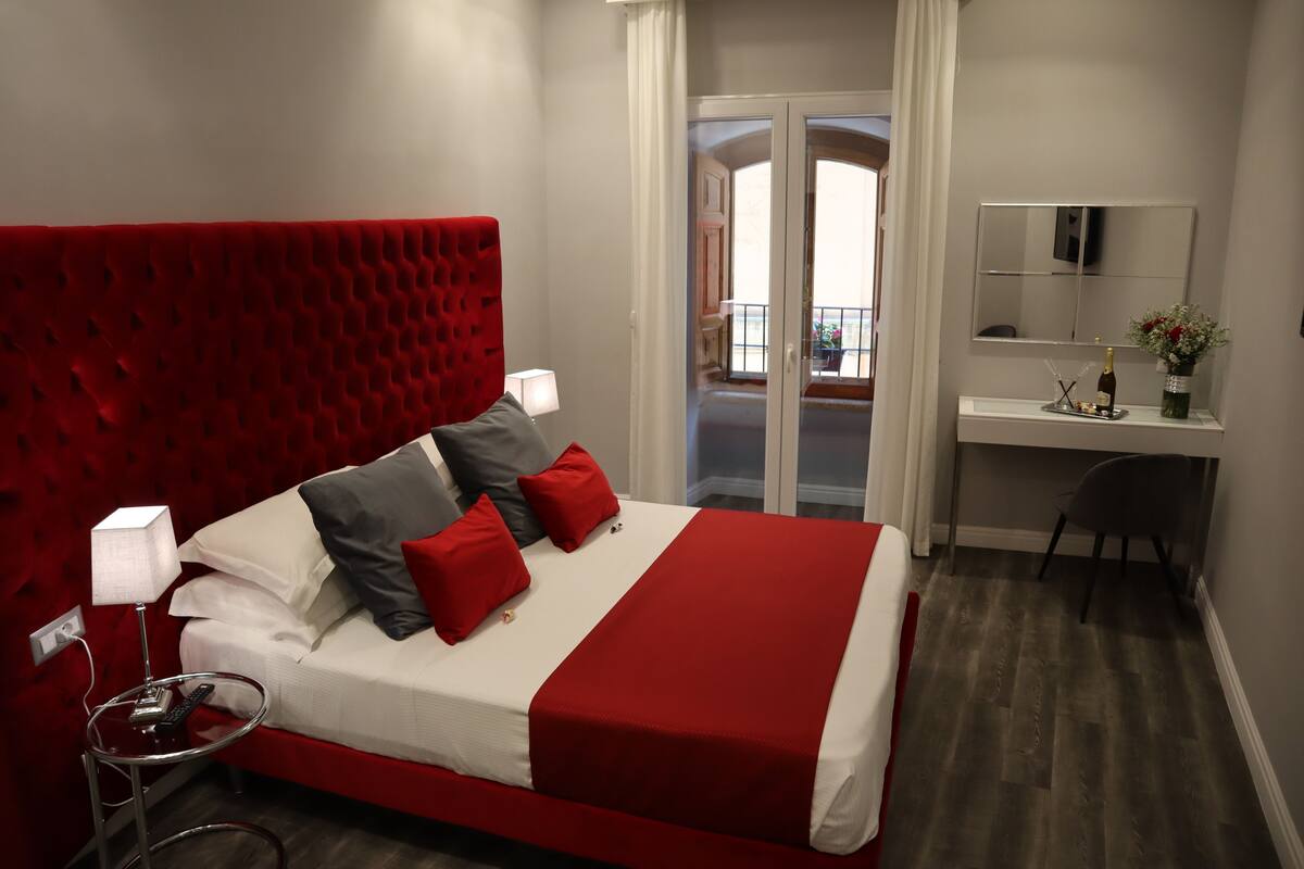 Quarto de hotel com cabeceira estofada em vermelho, colcha branca e almofadas vermelhas e cinzentas. Há uma mesa de trabalho com um espelho, uma garrafa de champanhe e um vaso com flores. Uma cadeira é visível no canto.
