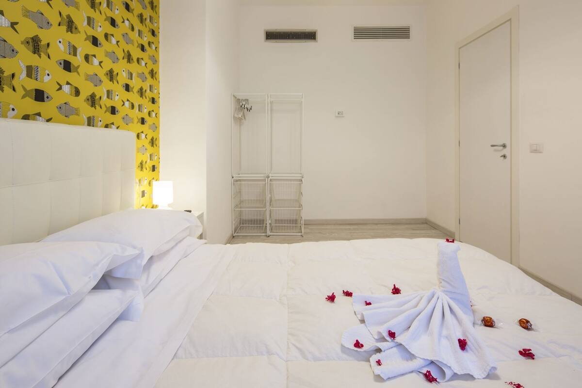 Quarto de hotel com cama, guarda-roupa, área de chuveiro e parede amarela com padrão de peixes.