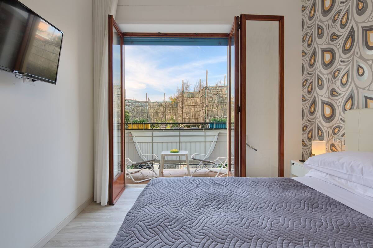 Quarto de hotel com cama, televisão, varanda com cadeiras e vista para prédios.