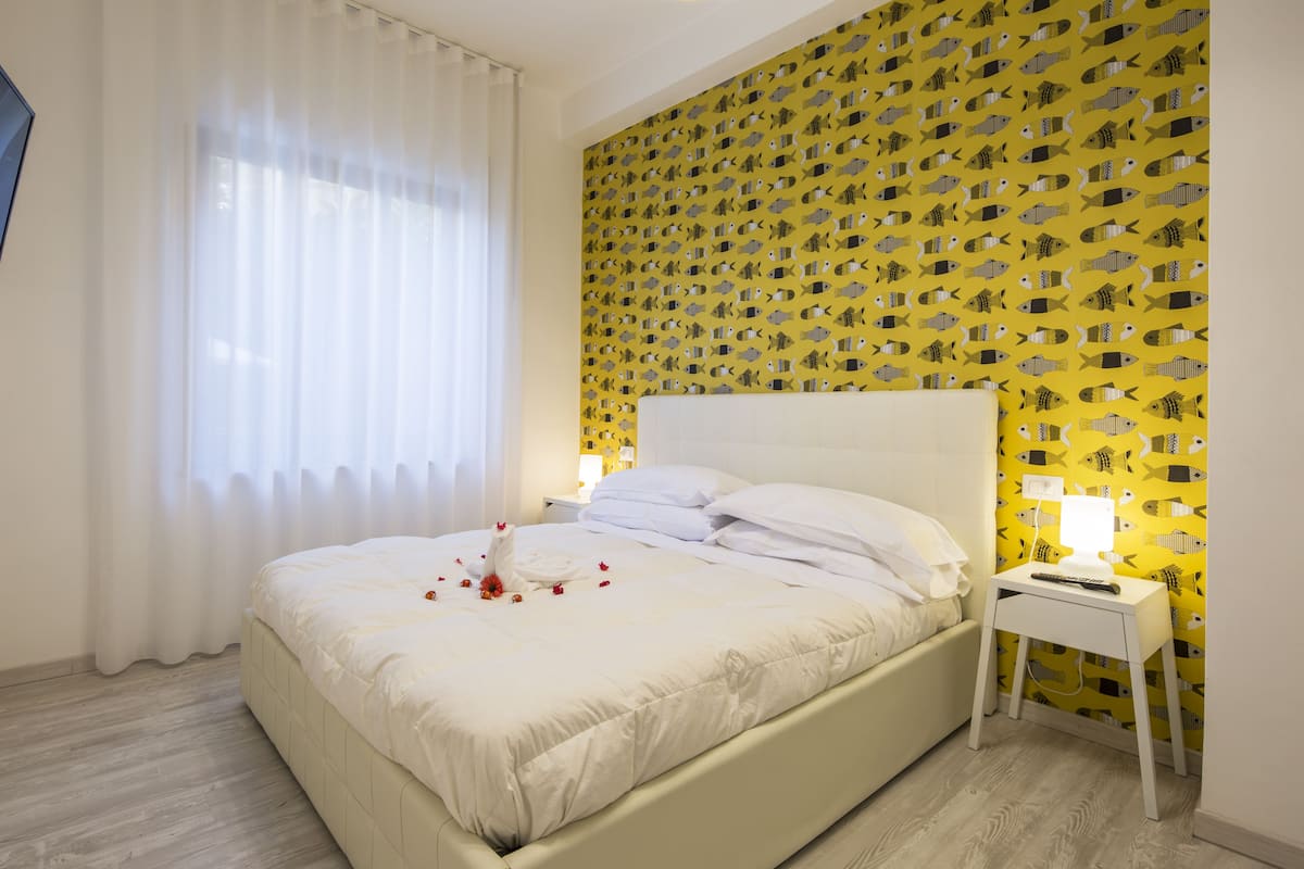Uma cama bem arrumada com lençóis e cabeceira brancos, uma mesa de cabeceira com abajur, e uma parede com padrão de peixes amarelos.