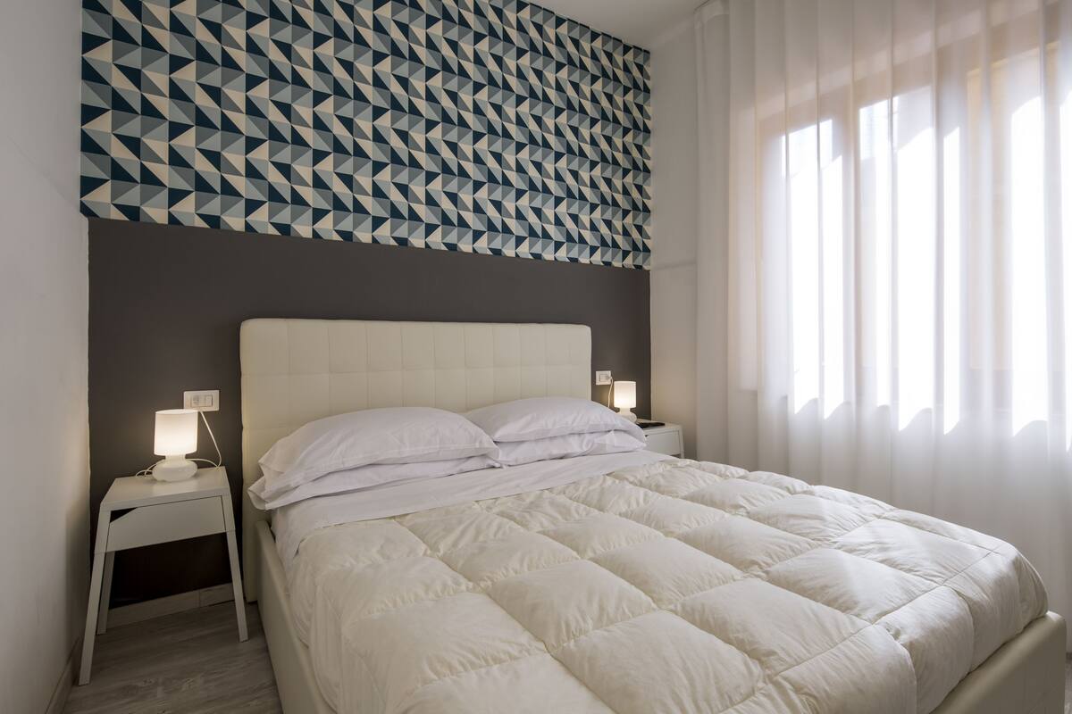 Quarto com parede estampada em padrão geométrico, cama branca com colcha, duas mesas de cabeceira com abajures e uma janela com cortinas leves.