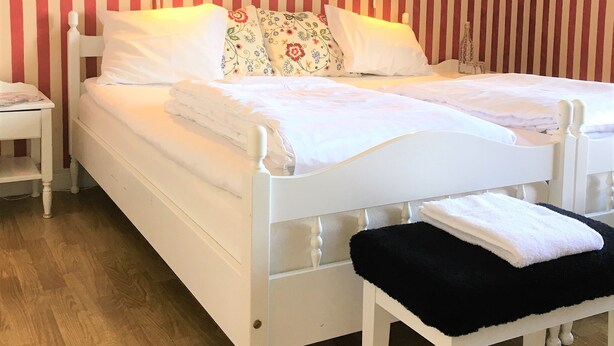 Uma cama de casal com roupa de cama branca e almofadas florais, uma mesinha de cabeceira com abajur, e um banquinho no rodapé da cama.