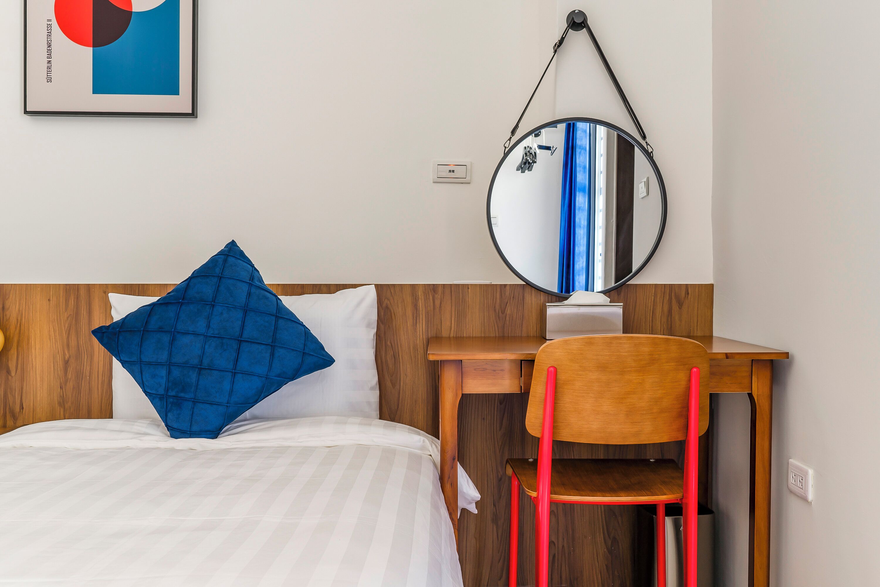 Quarto de hotel com cama, secretária de madeira, cadeira, espelho redondo e quadro emoldurado na parede.