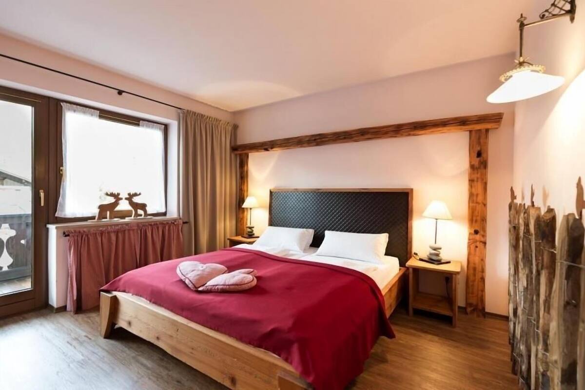 Quarto com cama de madeira, uma janela grande com cortinas, cabeceira e duas luminárias de cabeceira.