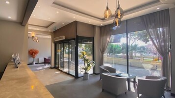 Um saguão moderno de hotel com balcão de recepção, área de estar, janelas amplas e vista para o exterior.