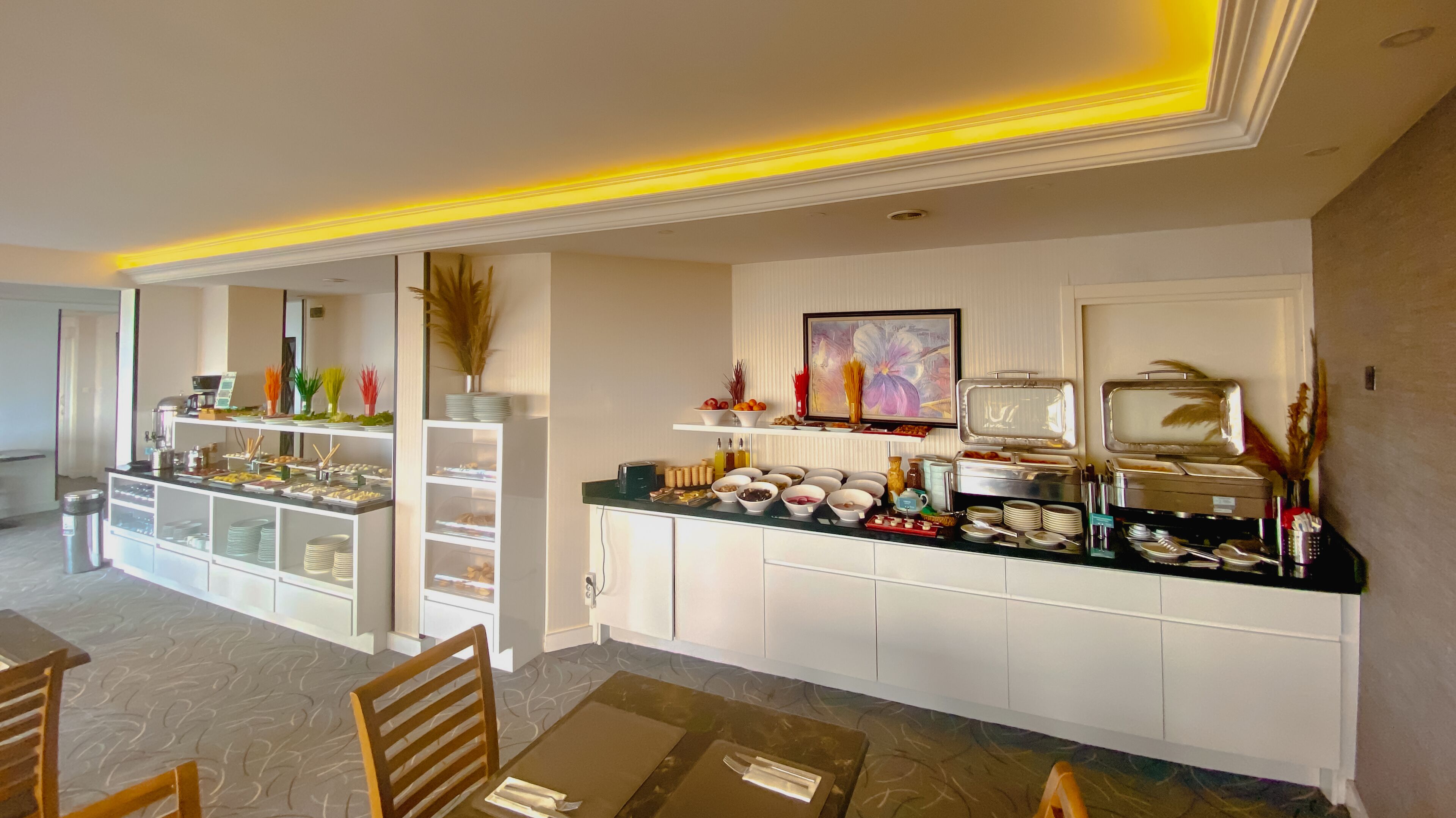Disposição de buffet com diversos alimentos e acompanhamentos, área de estar com cadeiras e um quadro decorativo na parede.