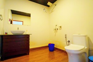Uma casa de banho com chão de madeira, lavatório branco, espelho, sanita e um balde azul.