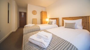 Quarto de hotel com duas camas, cada uma com cabeceira de madeira, roupa de cama branca e manta listrada. Há um guarda-roupa de madeira e uma luminária fixada na parede.