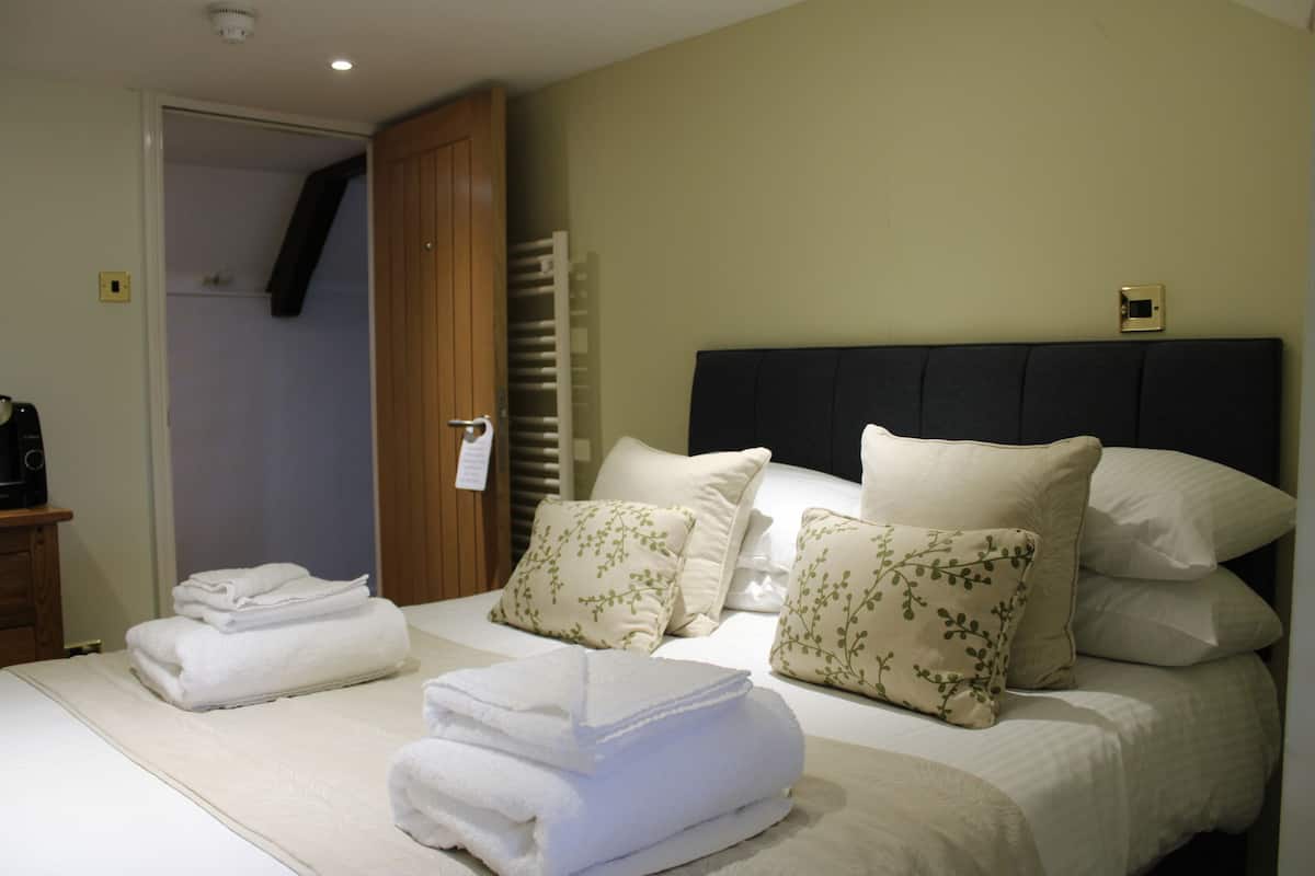 Cama bem arrumada com lençóis brancos e almofadas florais, porta de madeira e uma pequena mesa de cabeceira com abajur.