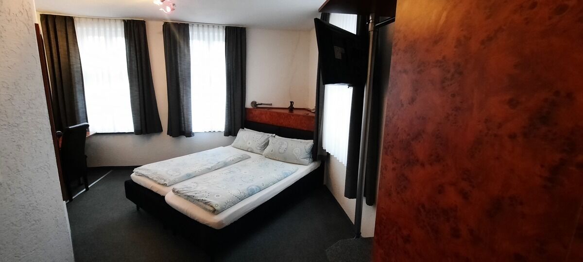 Quarto de hotel compacto com uma cama de solteiro, uma pequena mesa de trabalho e uma janela com cortinas.