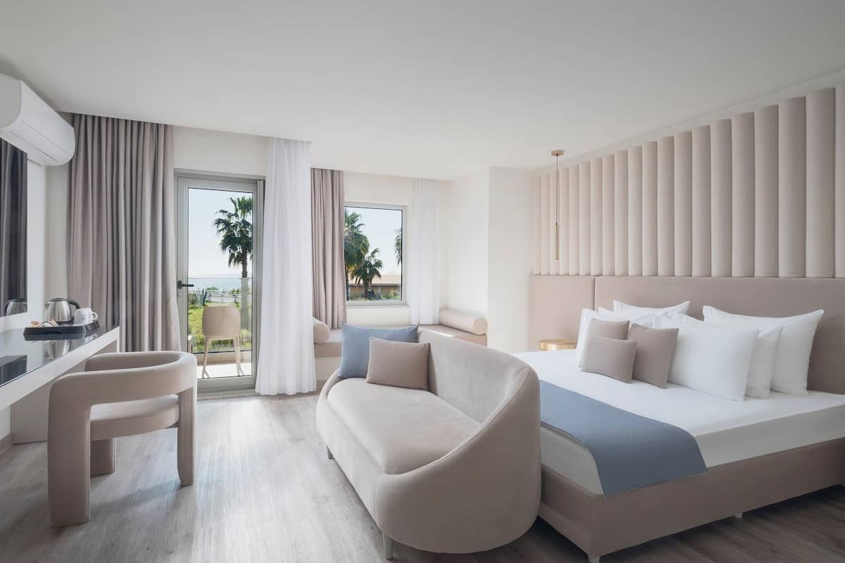 Quarto de hotel moderno com uma cama, sofá, escrivaninha e vista para palmeiras.