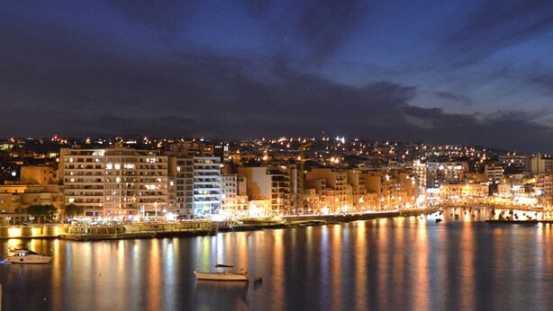 Paisagem urbana noturna com edifícios ao longo da orla, barcos na água e um rio calmo refletindo as luzes.