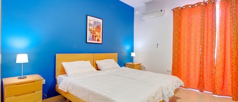 Quarto com cama, duas mesas de cabeceira, um criado-mudo, ar-condicionado fixado na parede e uma janela com cortinas laranja.