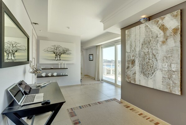 Uma sala de estar moderna com uma janela grande, uma escrivaninha com um laptop, uma pintura abstrata de grandes dimensões e um vaso decorativo com ramos.
