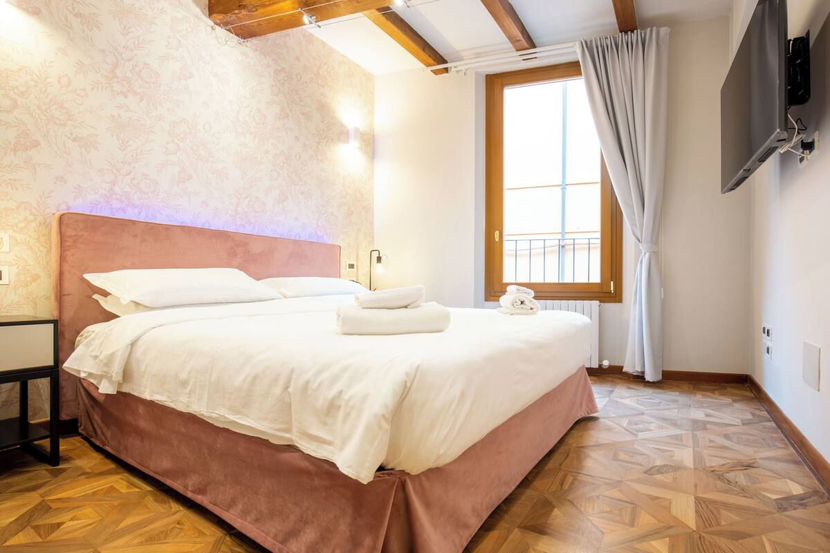 Cama bem arrumada com lençóis brancos, cabeceira de madeira e janela com cortinas.