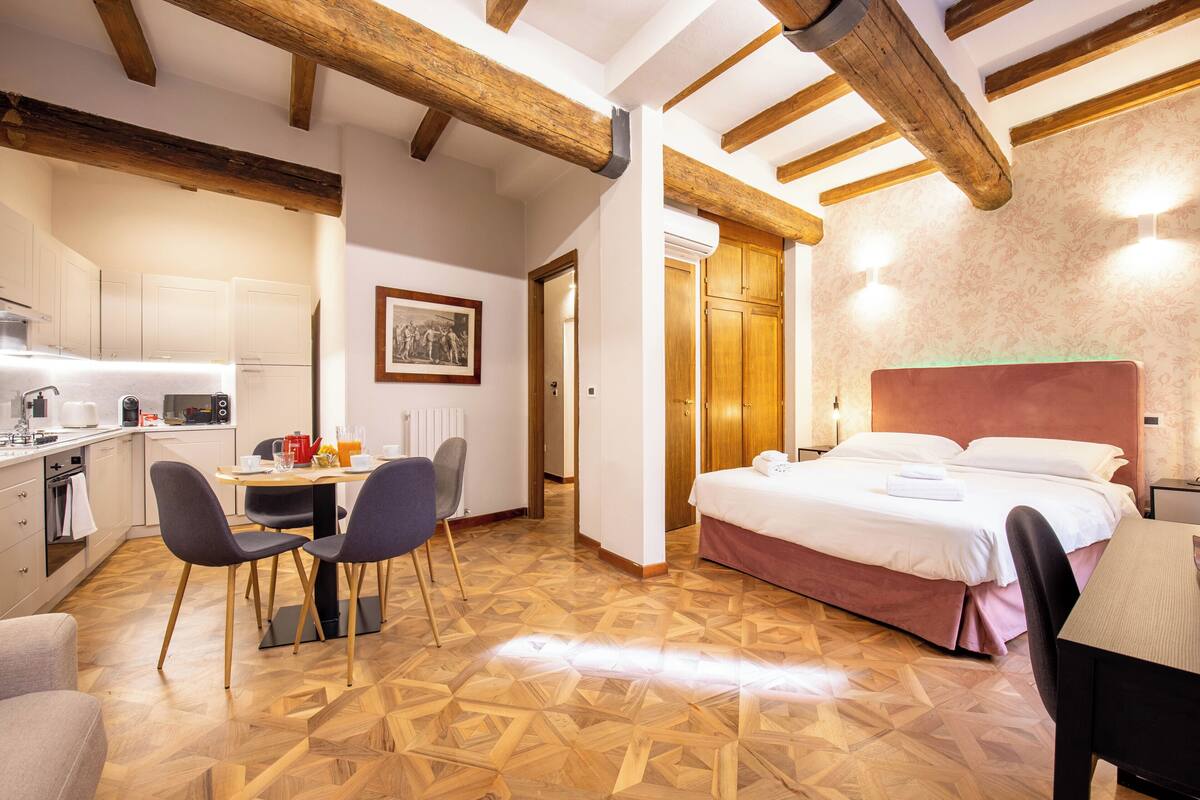 Quarto de hotel com teto de madeira, cama com roupa de cama branca, mesa de jantar com cadeiras e uma cozinha compacta.