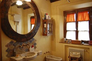 Casa de banho com um espelho redondo grande, uma pia com artigos de higiene, uma janela com cortinas laranja e um radiador.