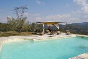 Piscina com espreguiçadeiras e toldo, cercada por árvores e montanhas.