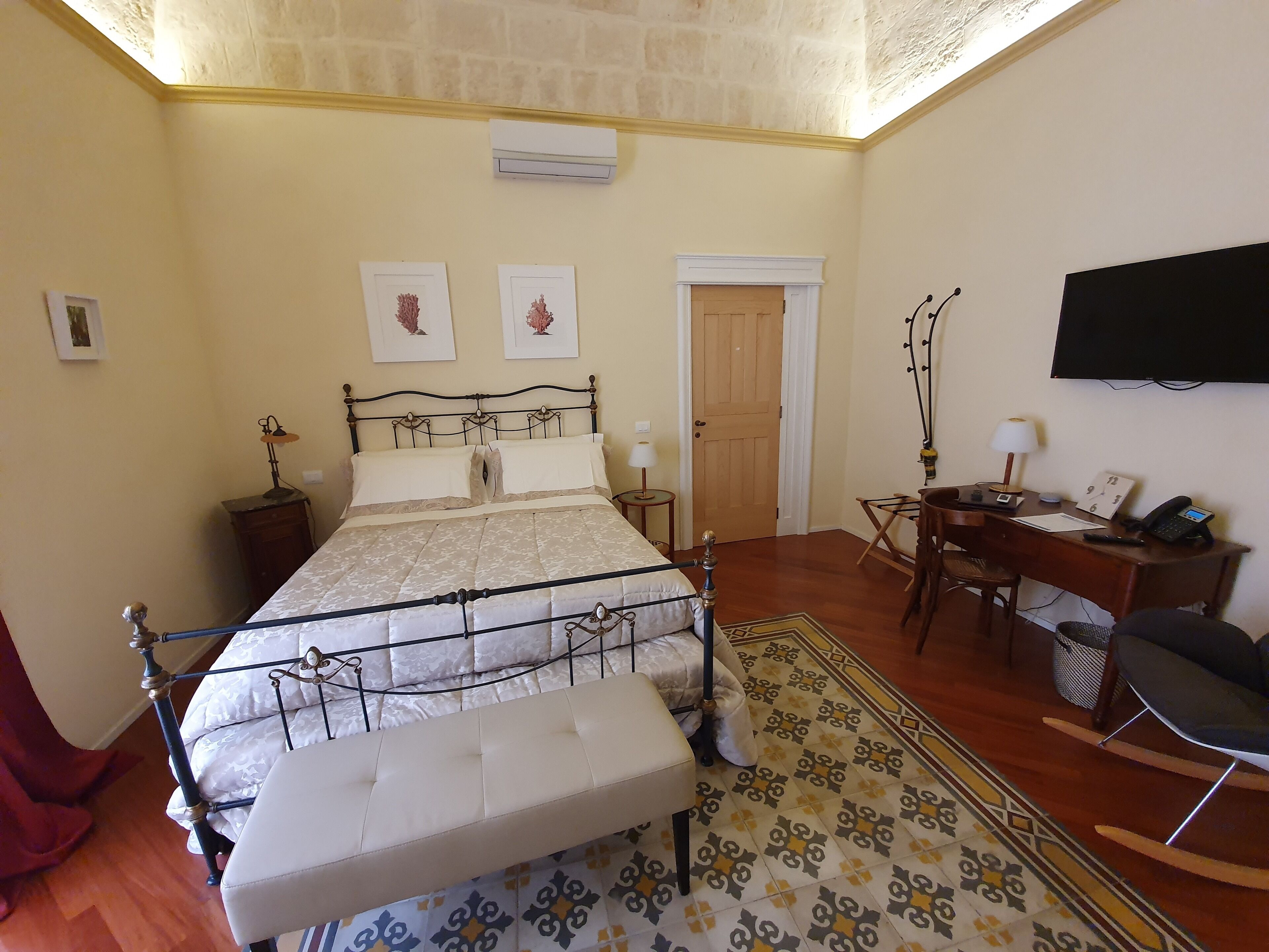 Quarto com cama, mesas de cabeceira, escrivaninha, cadeira, televisão e tapete estampado.
