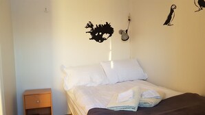Cama bem arrumada com lençóis brancos e toalha dobrada, mesa de cabeceira de madeira e luminária fixada na parede.