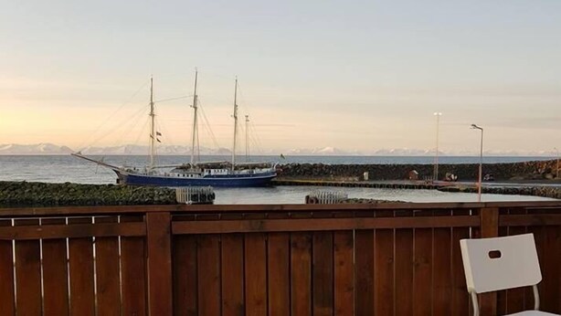 Um deck de madeira com uma mesa e uma cadeira, com vista para um corpo d'água onde se vê um veleiro e montanhas ao longe.