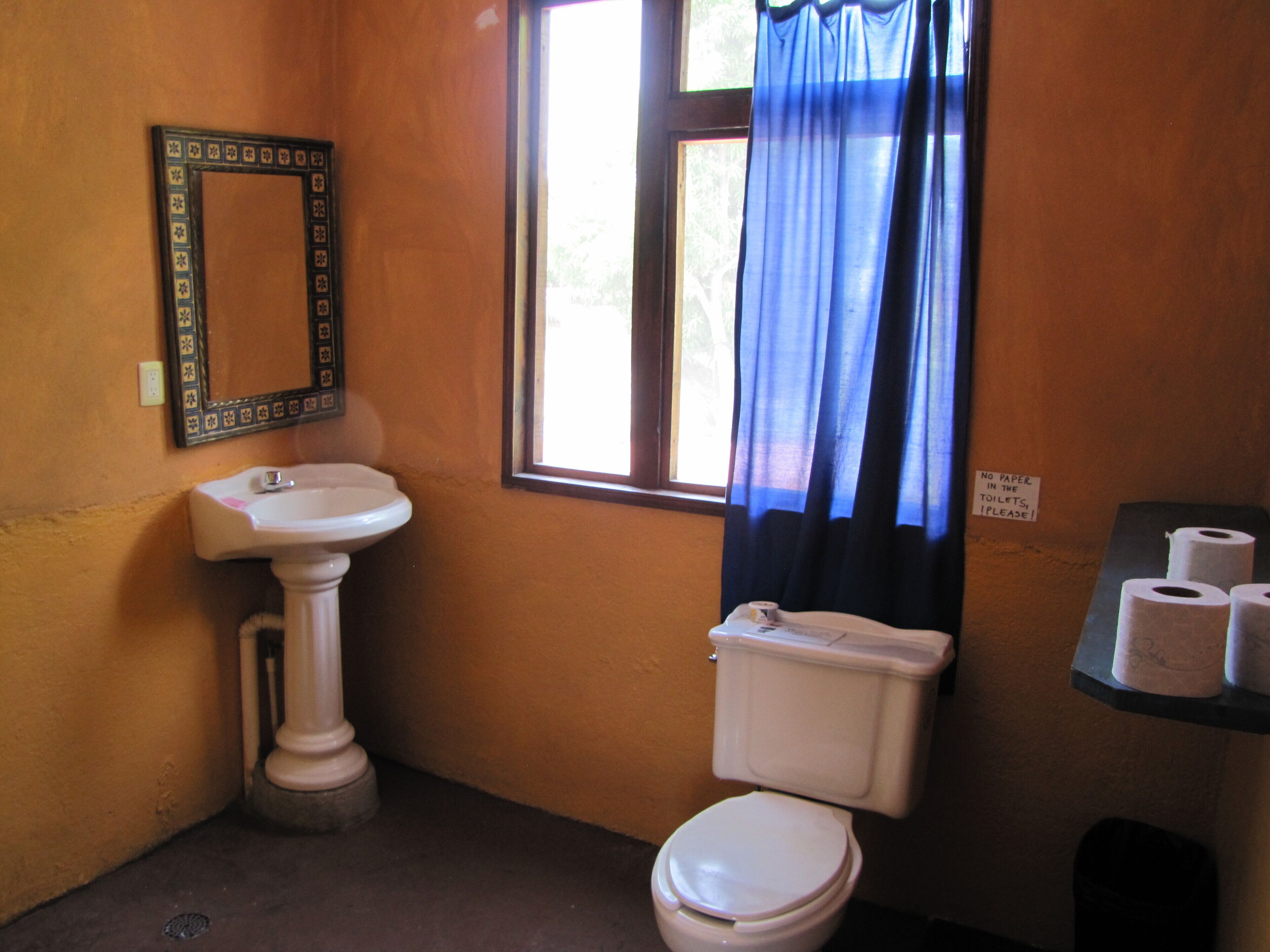 Casa de banho com lavatório assentado, sanita, janela com cortinas azuis e placa a proibir o uso de papel na sanita.