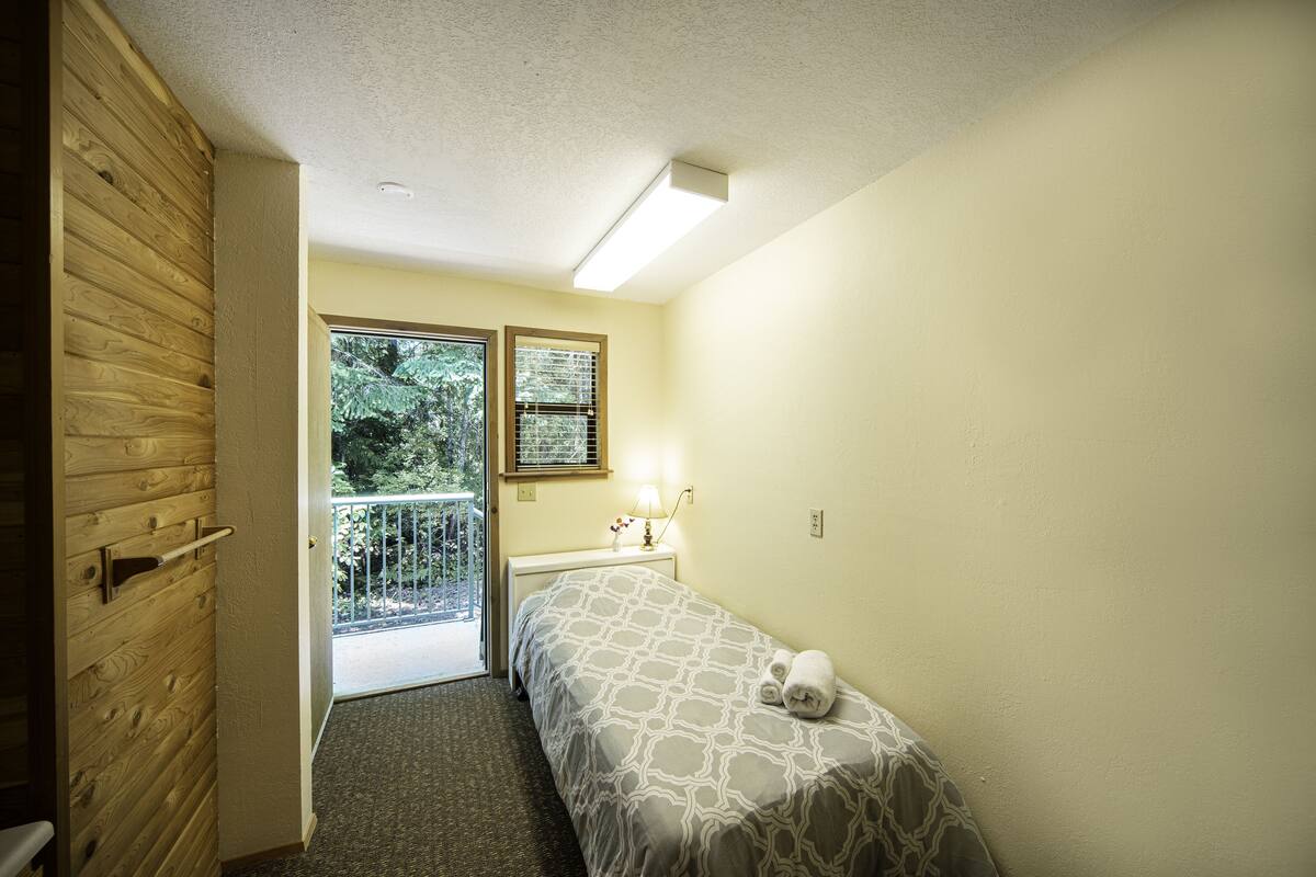 Quarto pequeno com uma cama de solteiro, porta de madeira, janela com persianas, mesa de cabeceira com abajur e vista para o exterior.
