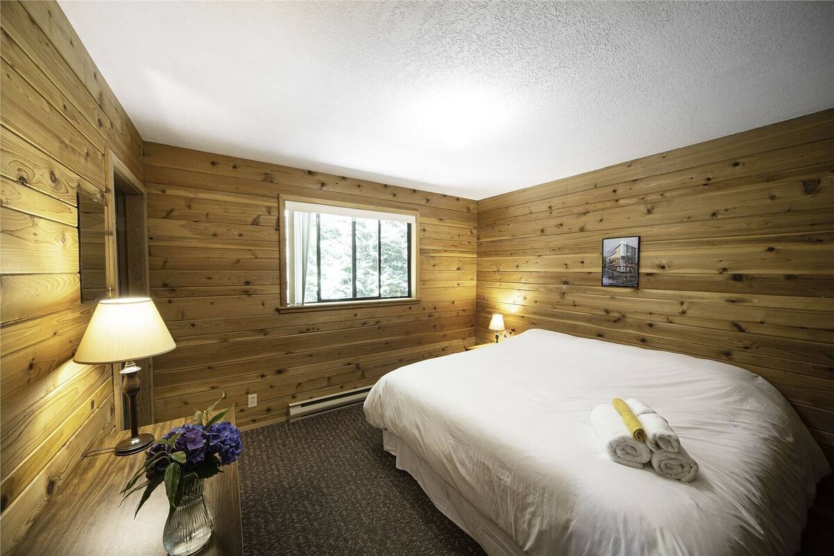 Quarto com painéis de madeira, uma cama, uma janela, um abajur e um quadro na parede.