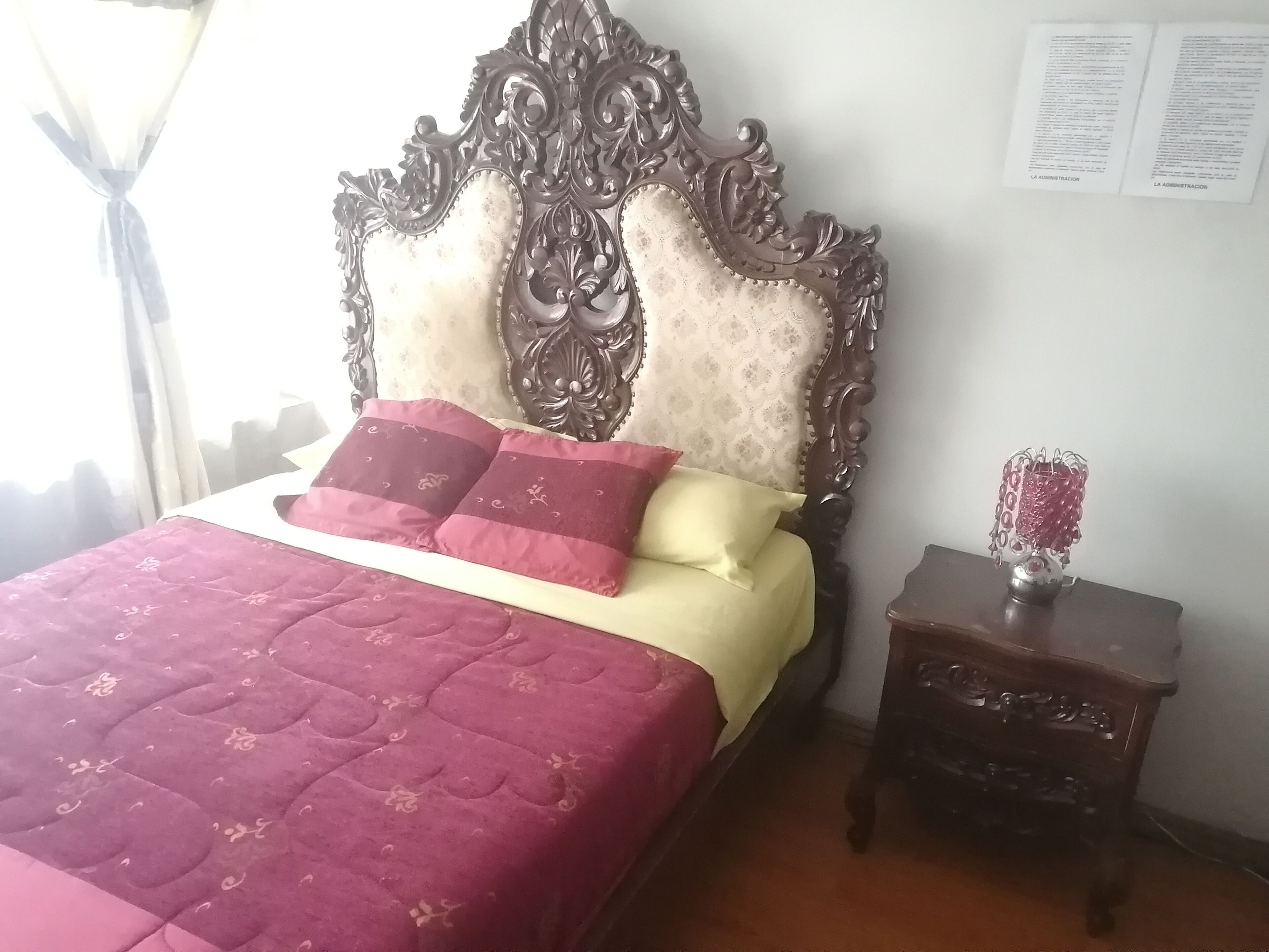 Uma cama bem arrumada com colcha estampada e almofadas, cabeceira de madeira, mesa de cabeceira com objeto decorativo e uma parede com um mural de avisos.