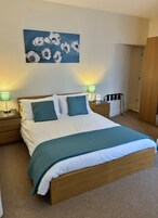 Cama bem arrumada com colcha azul e branca, duas almofadas combinando e um cobertor verde. Há duas mesas de cabeceira com luminárias, uma mesa de apoio de madeira e uma obra de arte na parede com flores brancas.