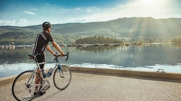 Uma pessoa andando de bicicleta em um caminho pavimentado, ao lado de um lago com barcos e colinas ao fundo.