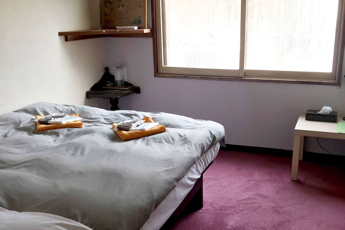 Um quarto com uma cama, uma mesinha, uma janela e uma estante.