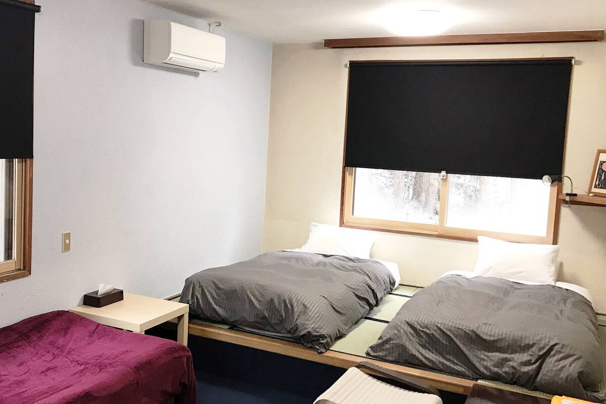 Quarto pequeno e limpo com duas camas, uma mesa de cabeceira, janela com cortina e ar-condicionado fixado na parede.
