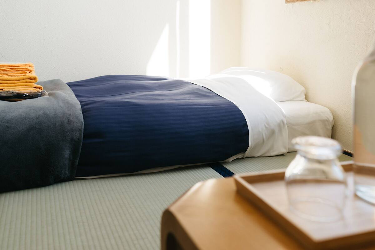 Uma cama bem arrumada com um edredom azul e branco, uma garrafa de vidro em um tabuleiro e uma pilha de toalhas dobradas em uma mesa de cabeceira.