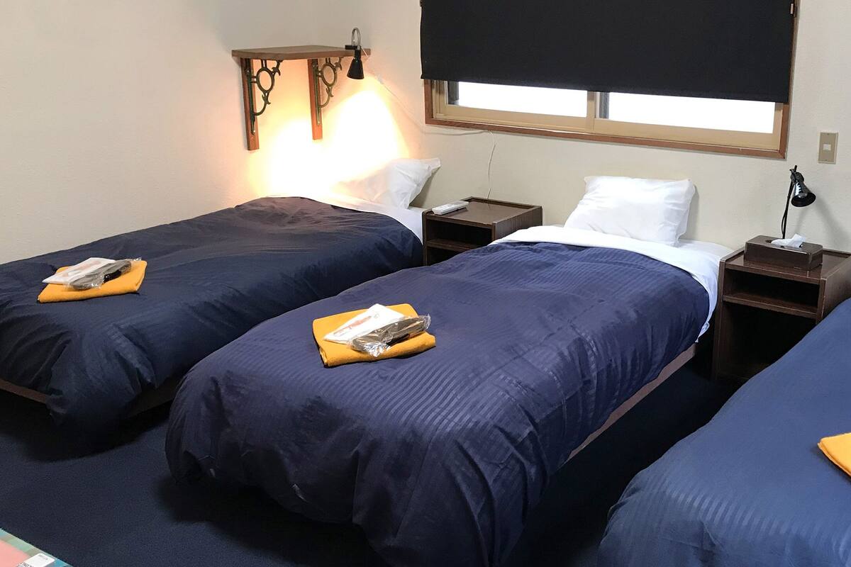 Quarto de hotel com três camas, cada uma com colcha azul e toalha amarela. Há mesas de cabeceira com luminárias e uma janela com persiana.