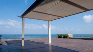 Terraço aberto com vista para o mar, com área de estar coberta e chão de tijolos.