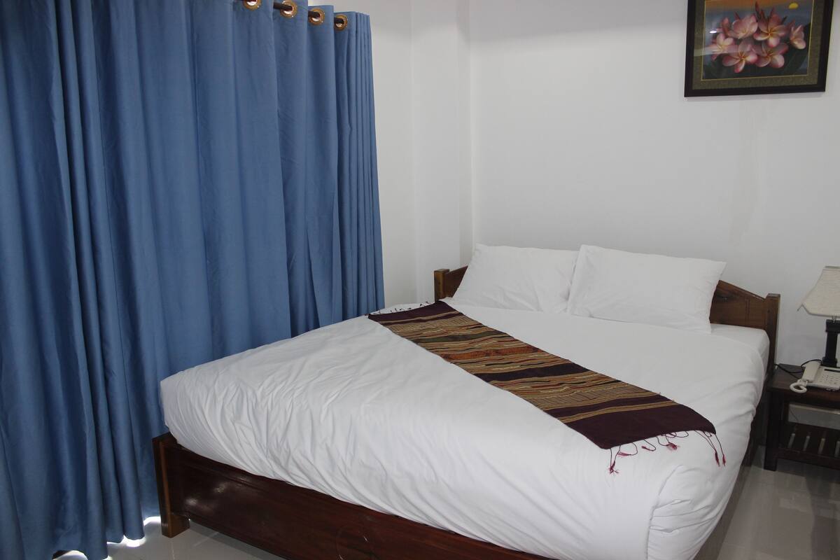 Uma cama bem arrumada com lençóis brancos e um cobertor estampado, cabeceira de madeira, mesa de cabeceira com telefone e cortinas azuis.