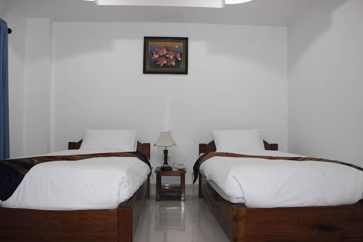 Quarto de hotel com duas camas de madeira, roupa de cama branca, uma pequena mesa de cabeceira com abajur e um quadro na parede.